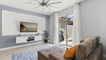 Uma sala de estar moderna com televisão de tela plana, ventilador de teto, porta de vidro deslizante para uma varanda e um sofá marrom com almofadas amarelas.