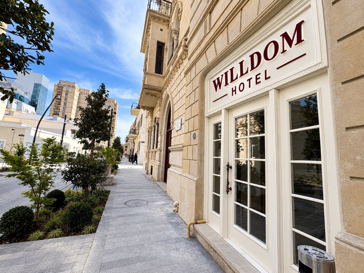 Exterior do Hotel Willdom, com vista clara da entrada, fachada de pedra e calçada arborizada.