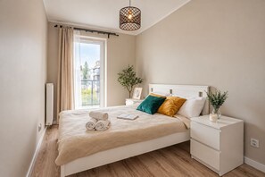 Quarto com cama, mesas de cabeceira, radiador, janela com cortinas e luminária pendente.