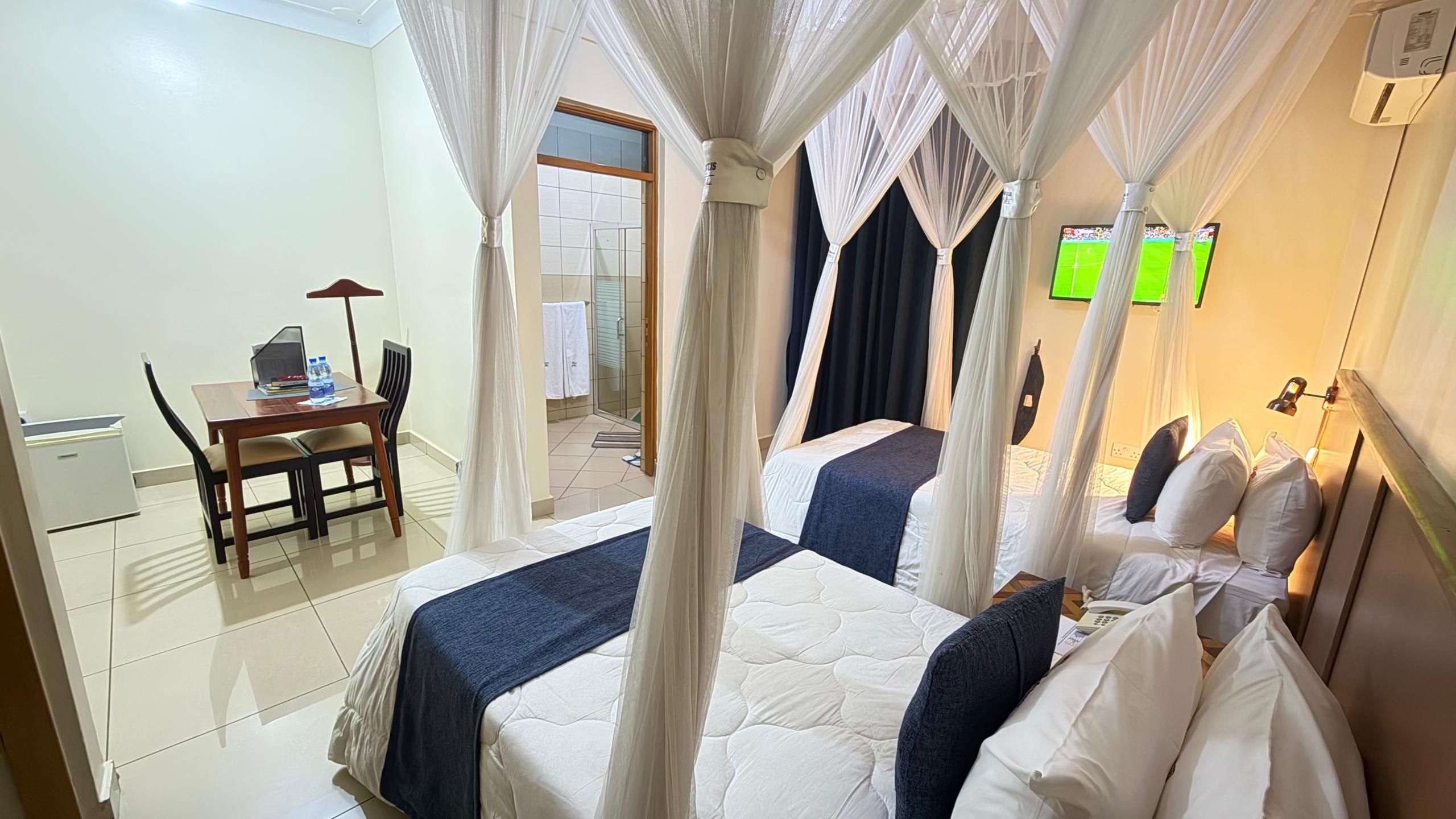 Quarto de hotel com duas camas, uma escrivaninha, uma cadeira, televisão e banheiro visível por uma porta aberta.