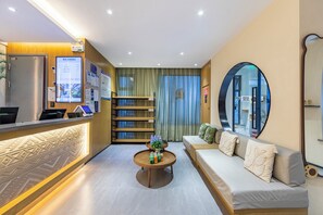 Uma área de recepção de hotel com balcão, espaço de espera com sofá e mesa de centro, e um espelho grande na parede.