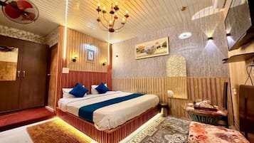 Quarto de hotel com uma cama grande, cabeceira de madeira, lustre, televisão e tapete estampado.