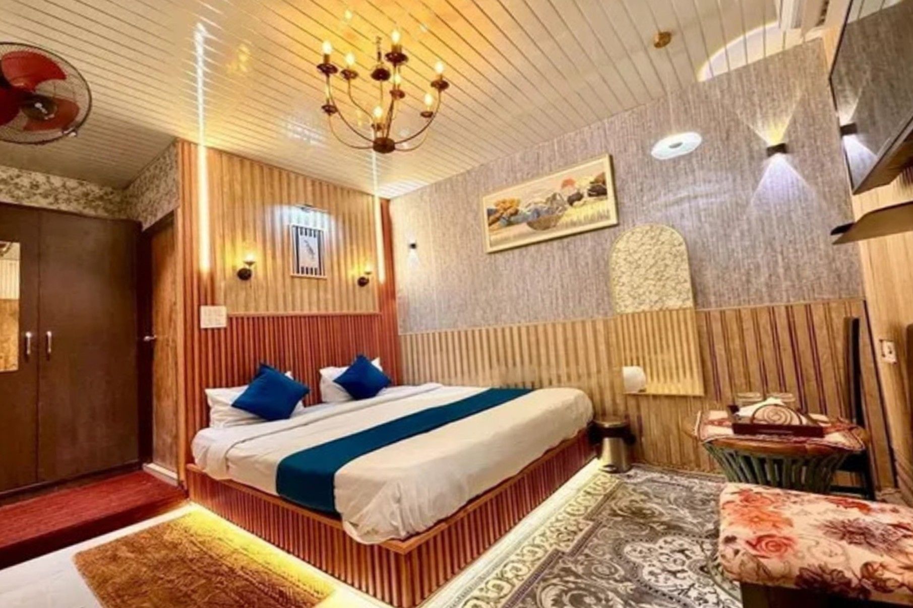 Quarto de hotel com uma cama grande, uma mesa pequena, uma cadeira, um ventilador, um relógio e um quadro na parede.