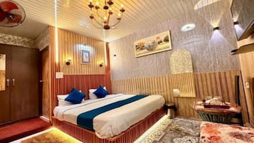 Quarto de hotel com uma cama grande, uma mesa pequena, uma cadeira, um ventilador, um relógio e um quadro na parede.