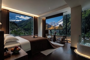 Quarto de hotel moderno com uma cama grande, área de estar e vista para montanhas e floresta.