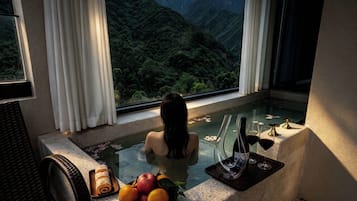 Pessoa em banheira com vista para montanhas, bandeja com frutas e copos de vinho.