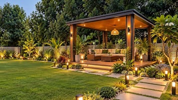 Área de estar coberta ao ar livre com sofá e mesa, cercada por um jardim com diversas plantas e um cercado de madeira.