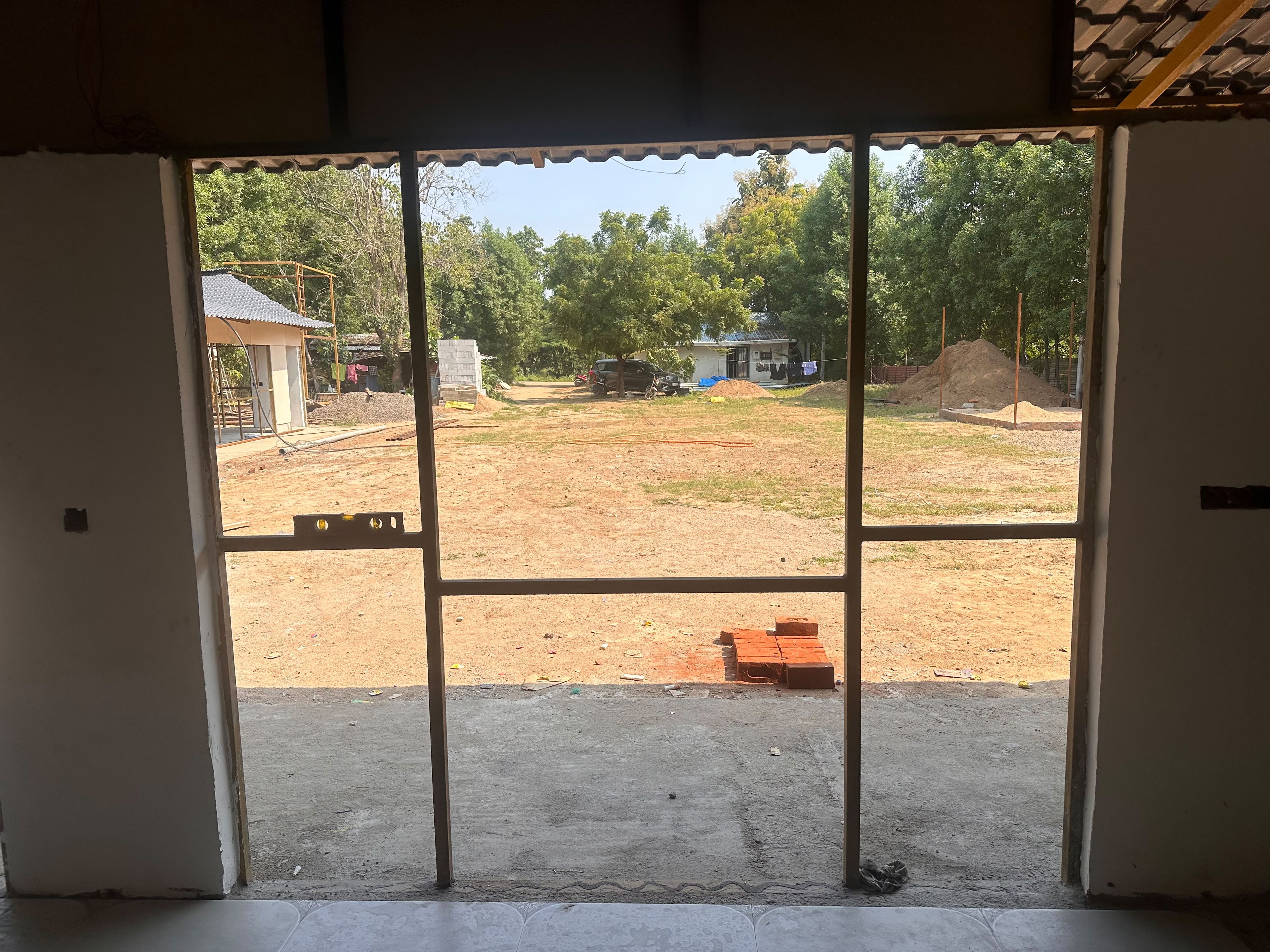 Visão pela janela com moldura de metal, mostrando uma área de construção com chão de terra, um muro de tijolos e algumas árvores.