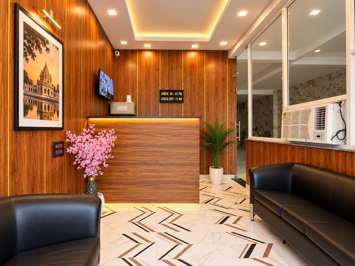 Uma área de recepção de hotel com um balcão de madeira, uma televisão, placa de check-in/out, uma planta em vaso e dois sofás de couro.