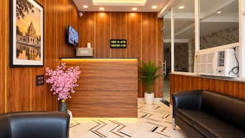 Uma área de recepção de hotel com um balcão de madeira, uma televisão, placa de check-in/out, uma planta em vaso e dois sofás de couro.