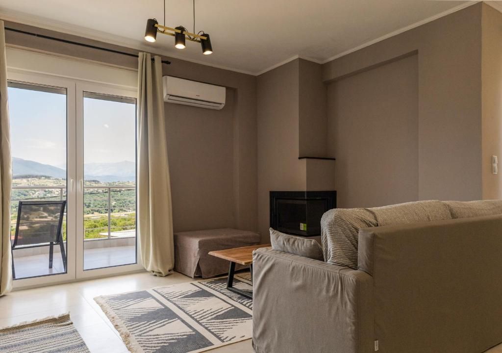 Sala de estar moderna com sofá, uma mesinha e televisão. Há uma porta de vidro deslizante que dá para uma varanda com vista para a paisagem.