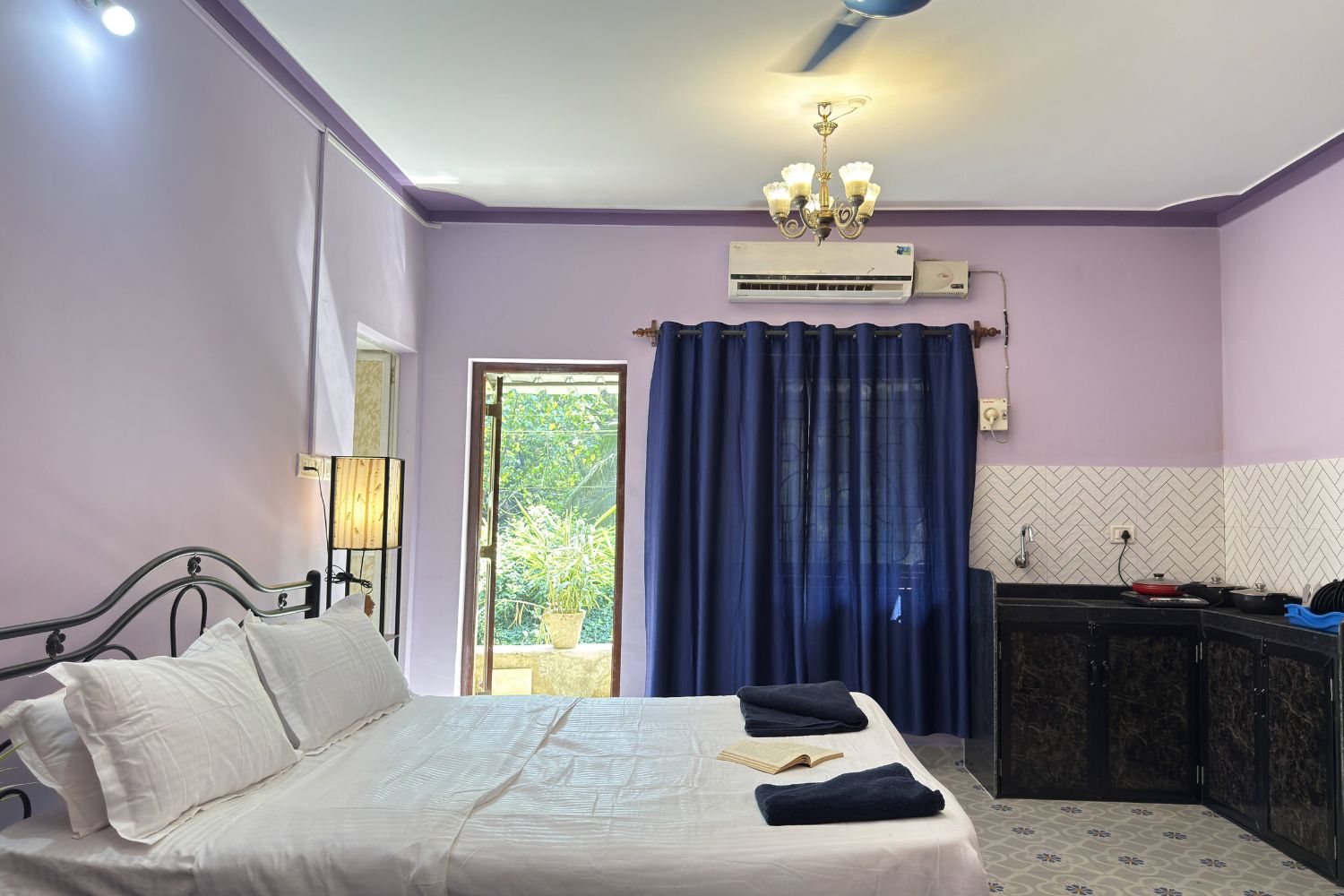 Quarto com cama, mesa de cabeceira, abajur, janela com cortinas, aparelho de ar condicionado e área de cozinha com pia e bancada.