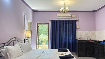 Quarto com cama, mesa de cabeceira, abajur, janela com cortinas, aparelho de ar condicionado e área de cozinha com pia e bancada.