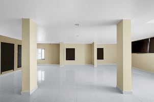 Um quarto amplo e vazio, com piso de azulejos brancos e paredes bege. Possui três pilares grandes e várias janelas.