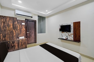 Quarto de hotel com cama, guarda-roupa, televisão na parede e uma pequena prateleira com objetos.