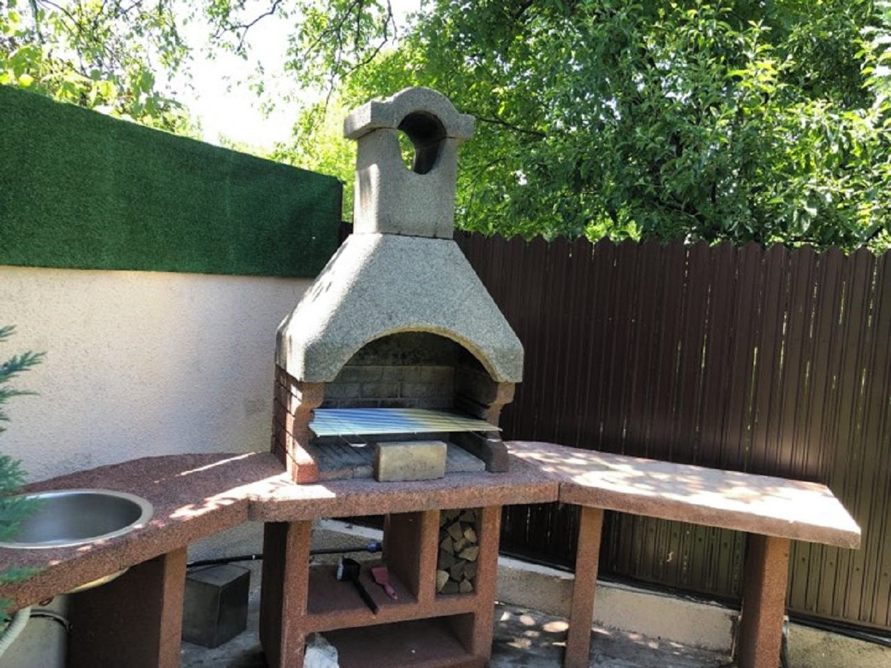 Forno de pizza de pedra ao ar livre, com base de tijolos e chaminé, cercado por uma mesa de madeira e uma pia de metal.