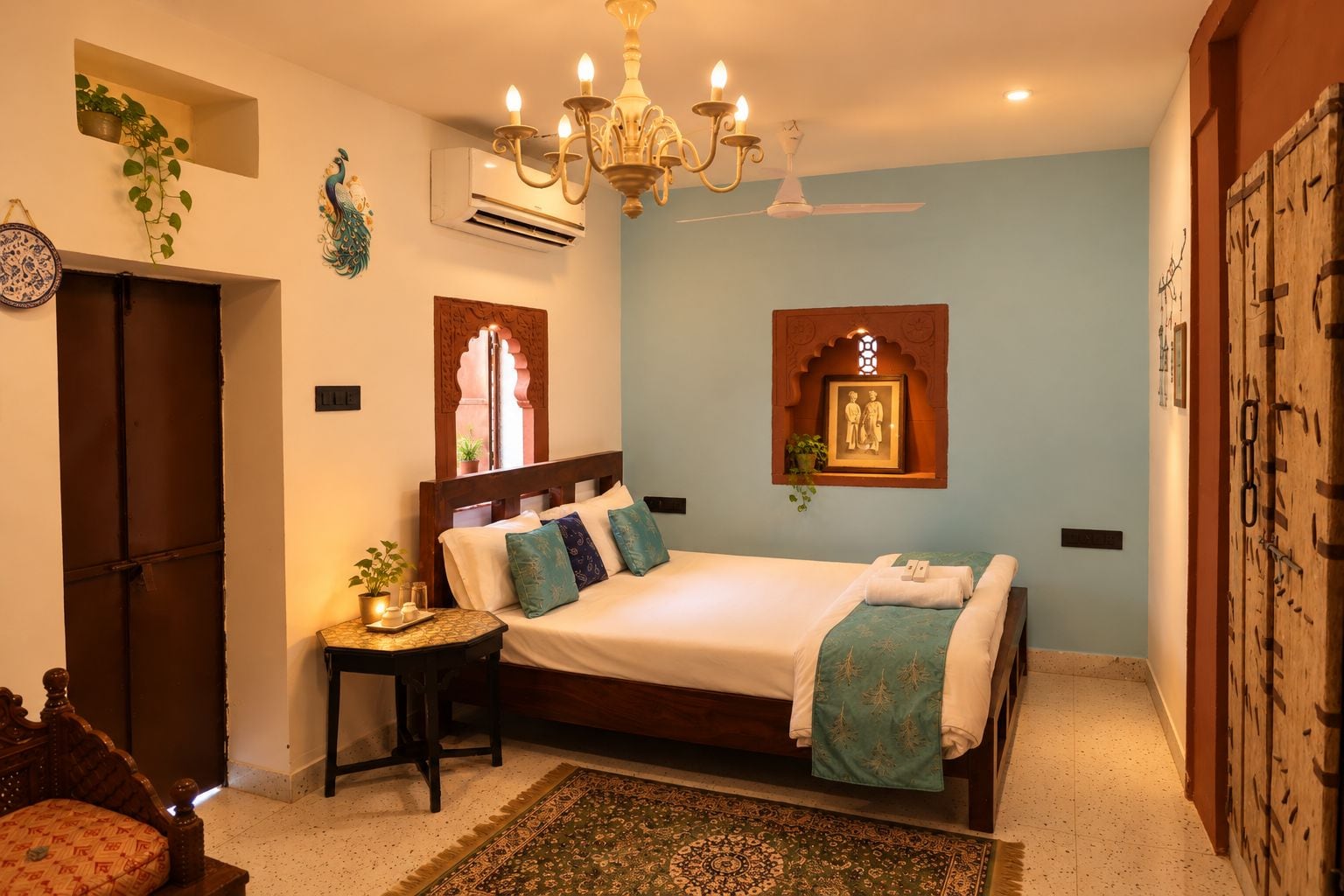 Quarto com cama, mesa de cabeceira, lustre, ar-condicionado e porta com painel decorativo.