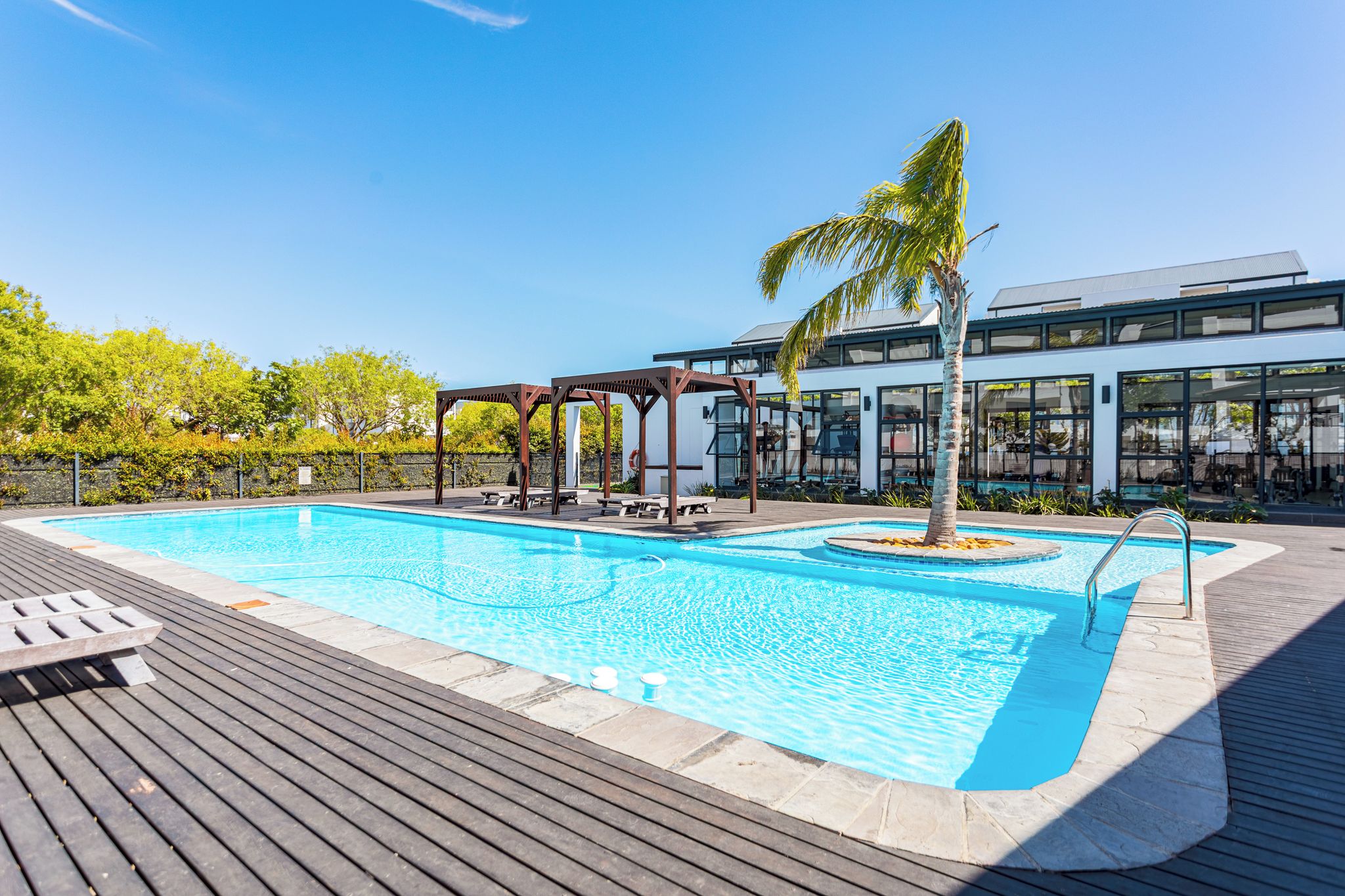 Uma piscina moderna ao ar livre com um deck de madeira, uma área de estar coberta e uma palmeira.