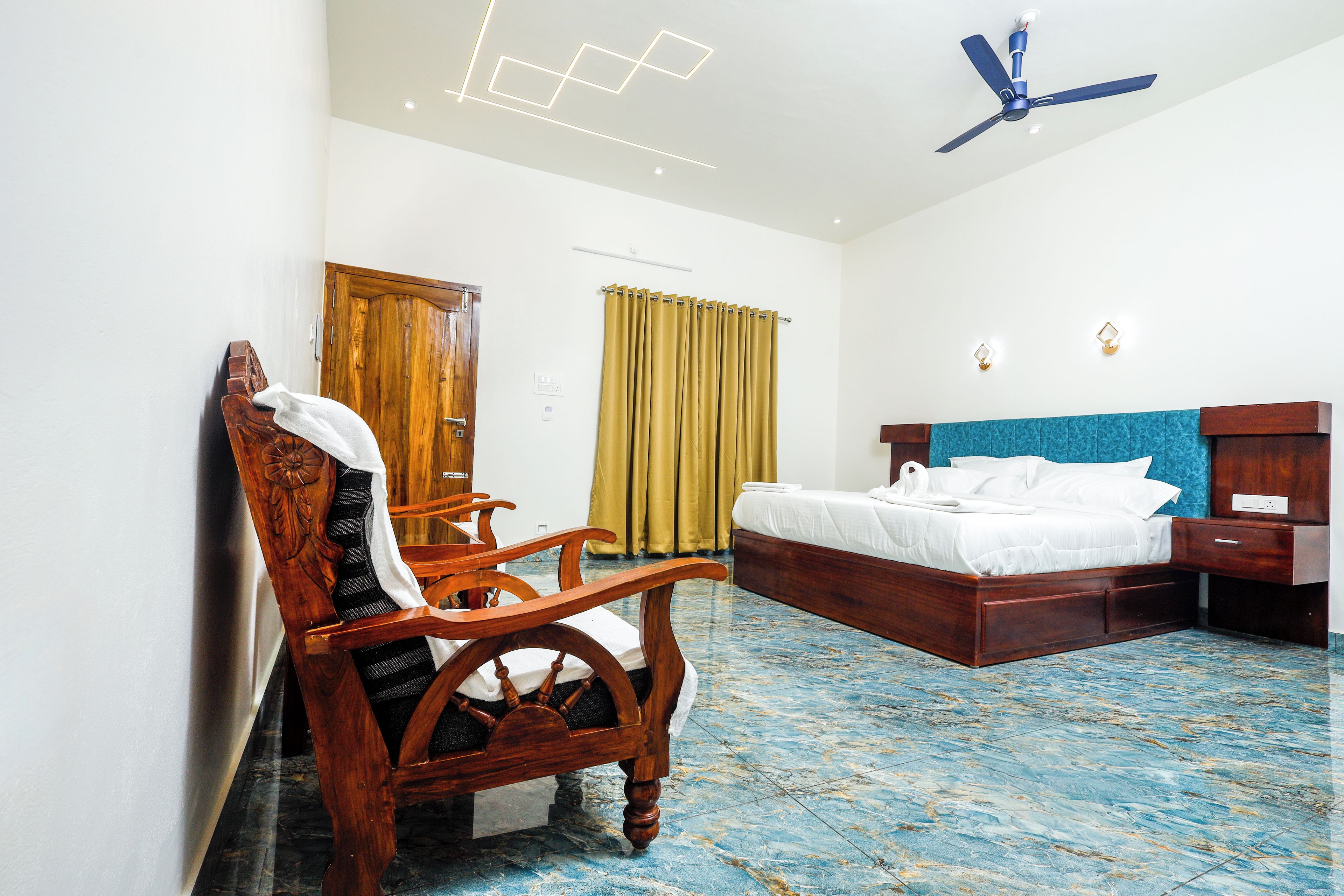 Quarto de hotel com cama, cadeira de madeira, escrivaninha, ventoinha no teto e piso azul estampado.