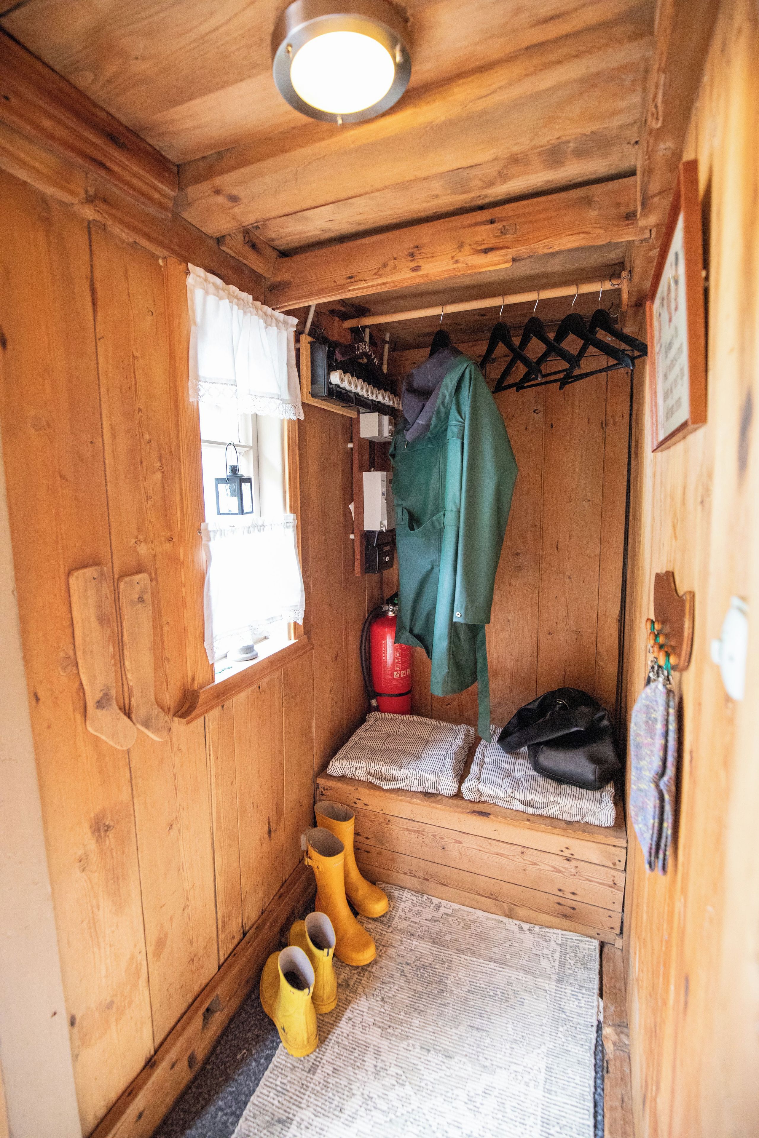 Uma cabana pequena com interior de madeira, uma janela com cortinas brancas, um cabideiro com um casaco verde, um extintor de incêndio e um par de botas amarelas.