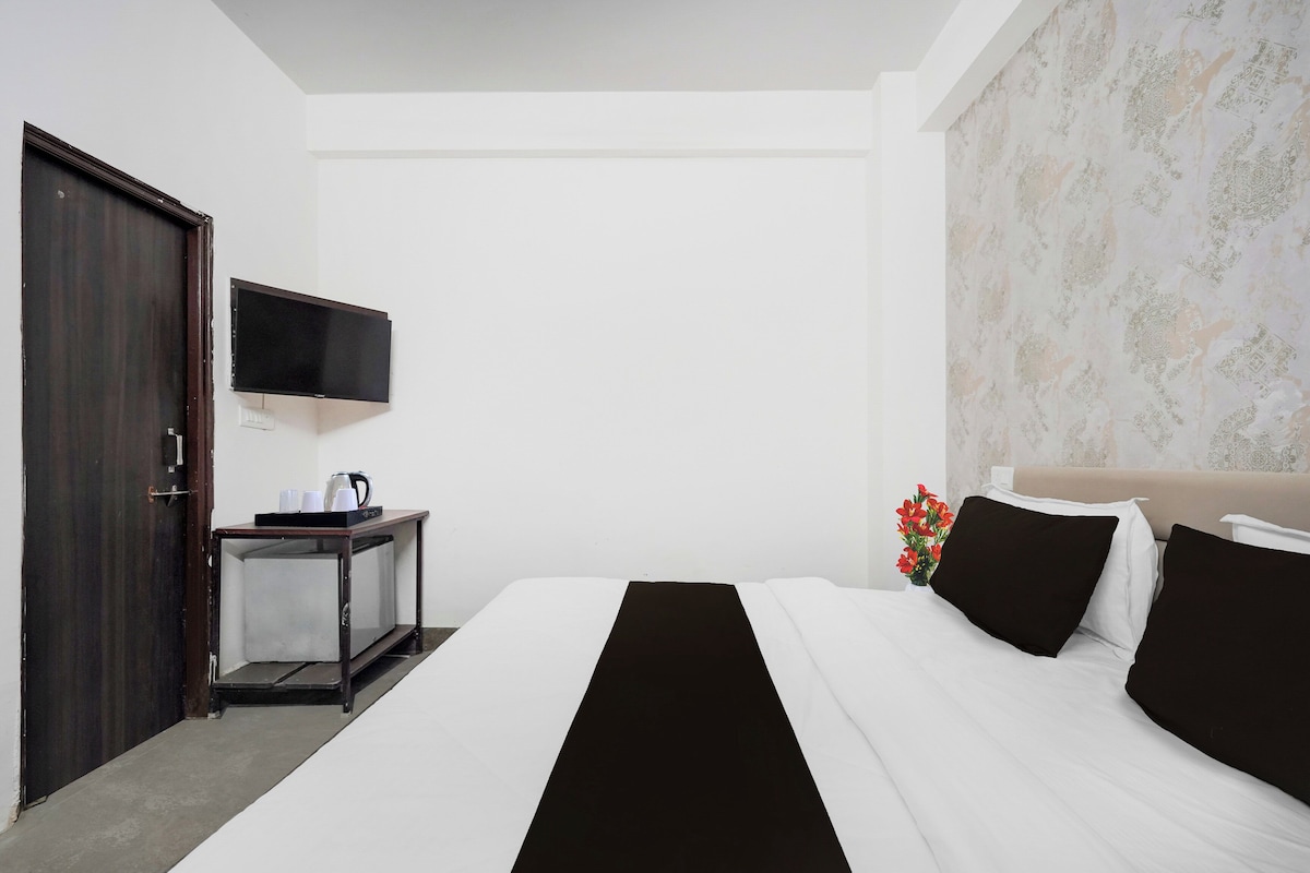 Quarto de hotel com uma cama, televisão presa na parede, uma pequena mesa com um tabuleiro de chávenas e uma chaleira, e uma porta.