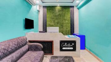 Uma área de recepção com balcão, sofá, TV fixada na parede e um painel de grama artificial verde.