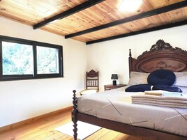 Quarto com teto de madeira, uma cama grande com cabeceira, uma janela e uma mesa de cabeceira com abajur.
