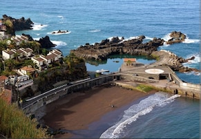 Uma área costeira com praia de areia, afloramentos rochosos e uma zona residencial com casas.