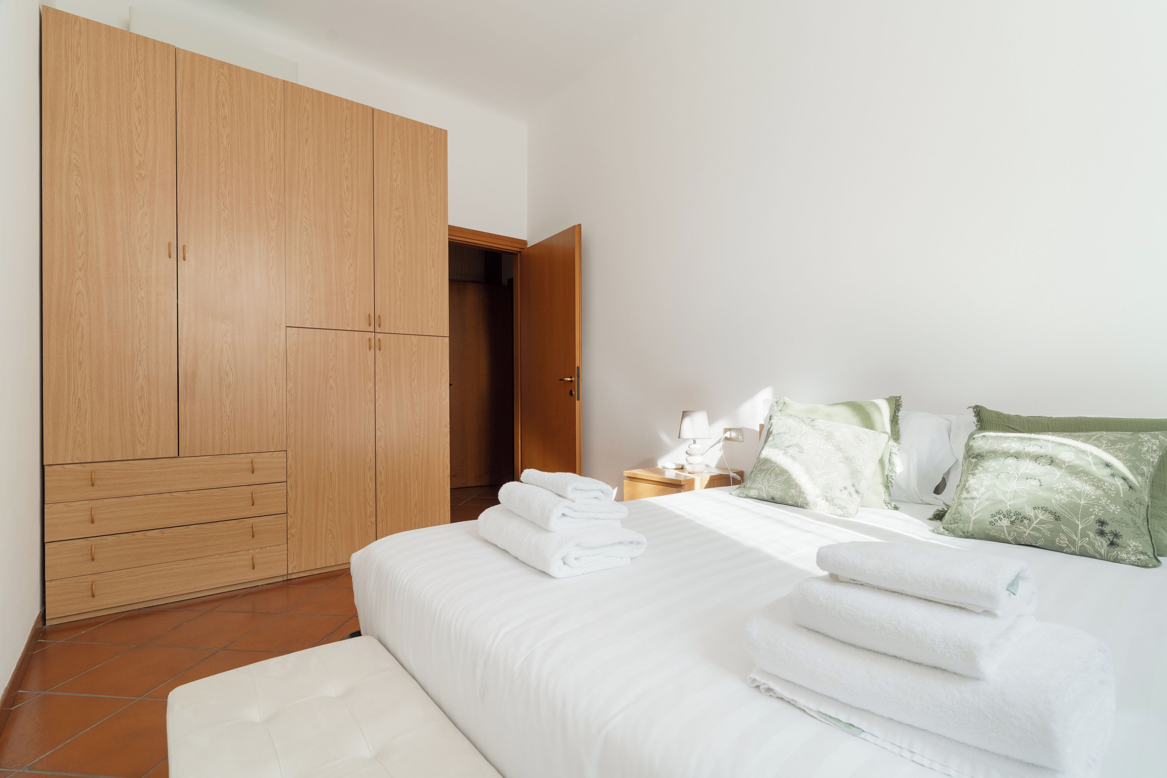 Quarto de hotel com um guarda-roupa espaçoso, cama bem arrumada com roupa de cama branca e mesa de cabeceira com abajur.