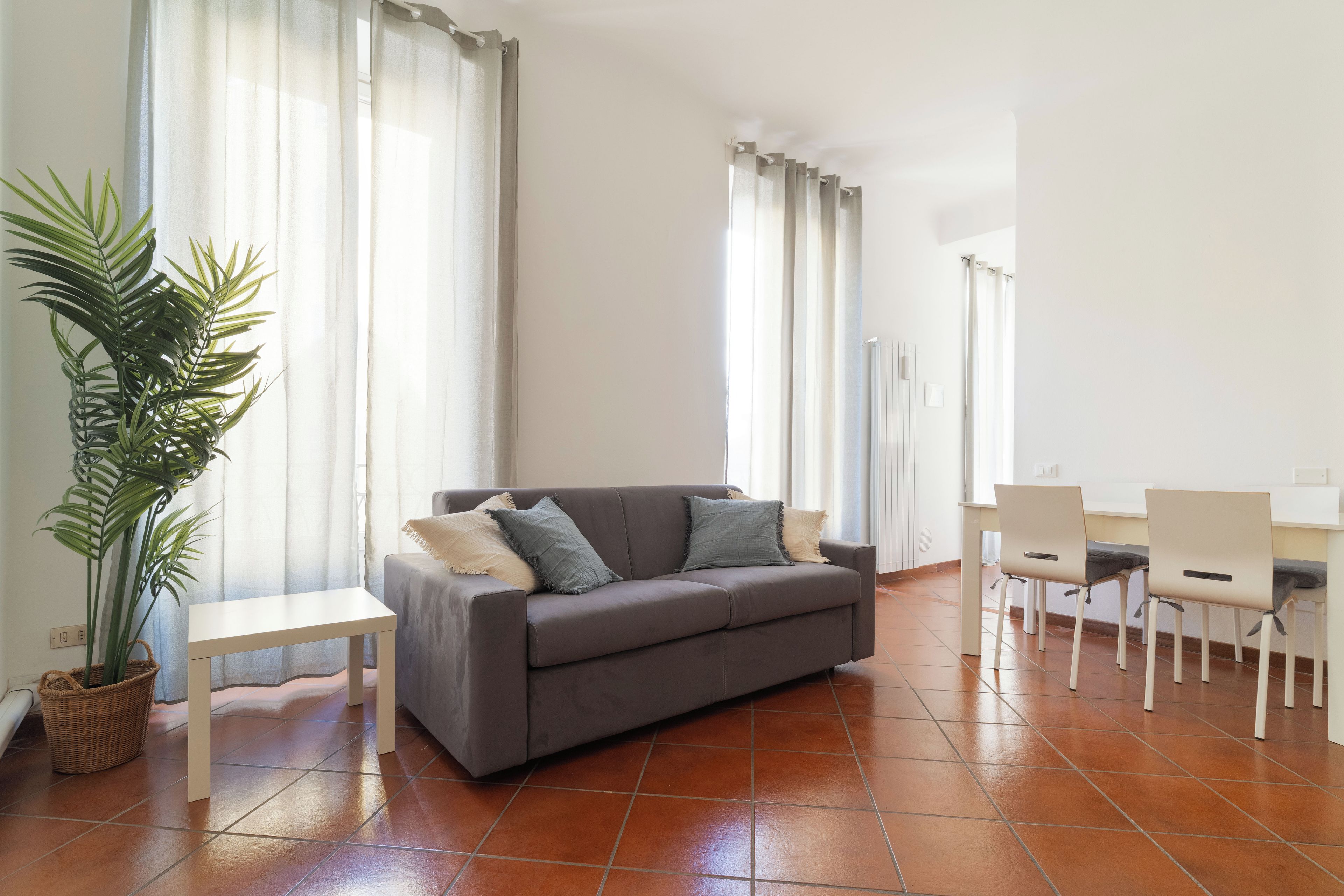 Uma sala de estar moderna com um sofá cinzento, duas poltronas, uma mesa de jantar com cadeiras e uma grande planta em vaso.