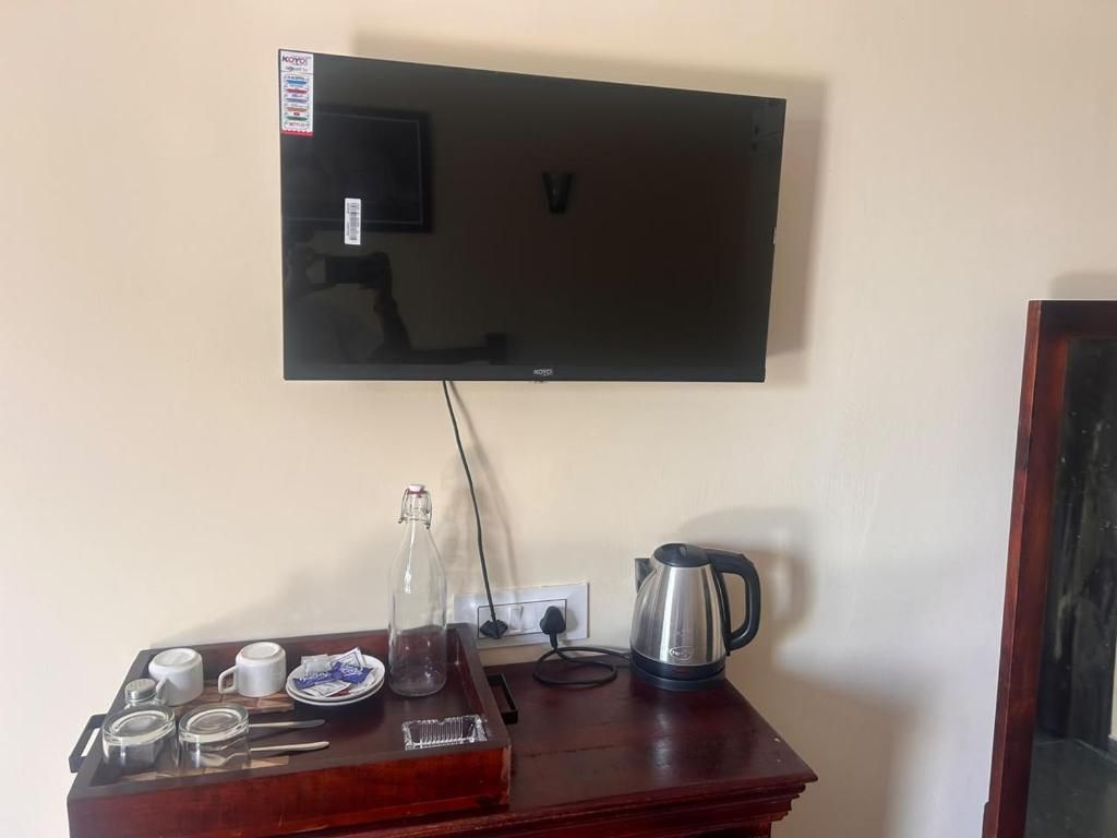 Televisão de tela plana fixada em parede, acima de uma mesa de madeira com cafeteira, xícaras e jarra de vidro.