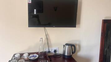 Televisão de tela plana fixada em parede, acima de uma mesa de madeira com cafeteira, xícaras e jarra de vidro.
