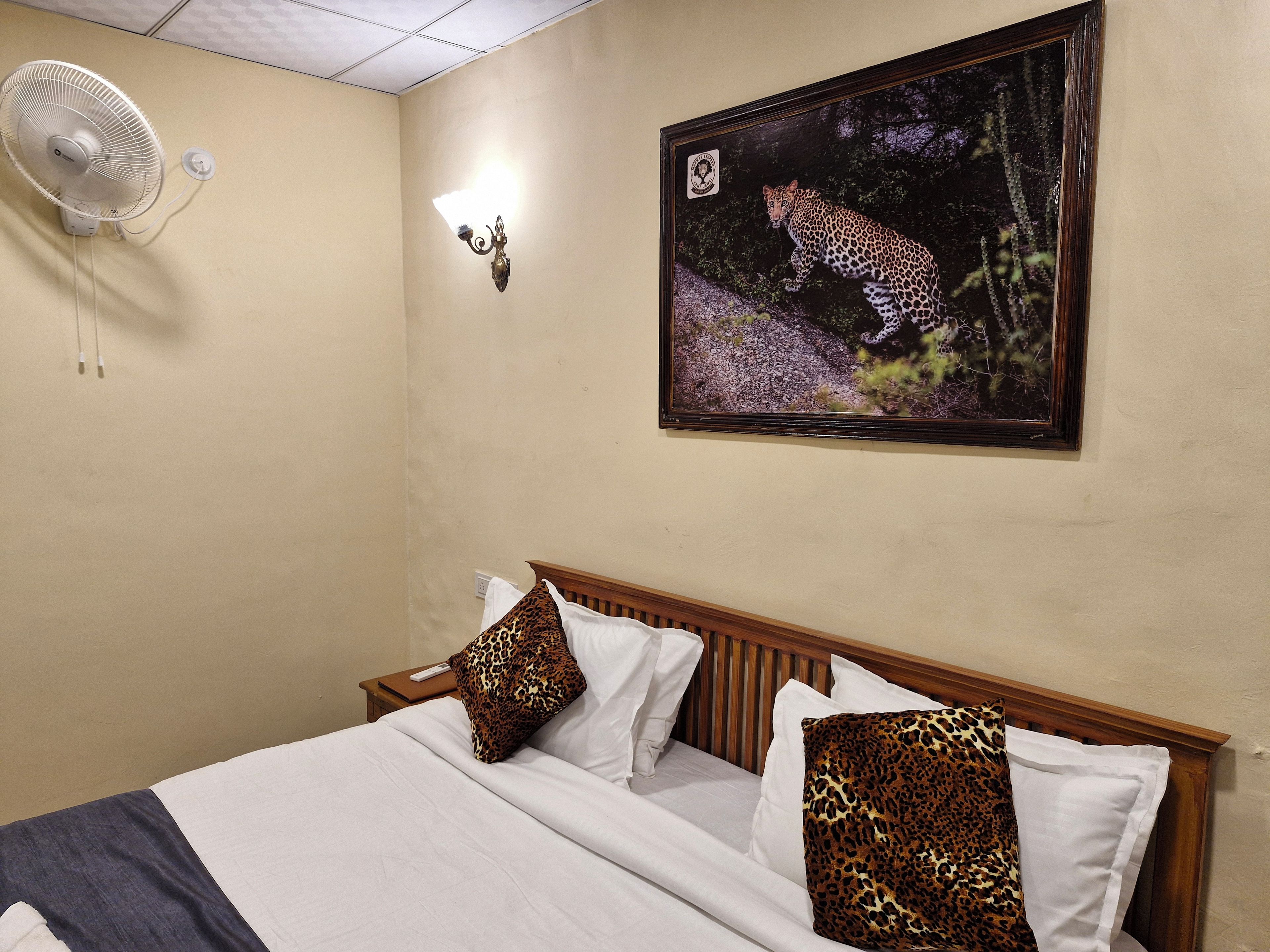 Quarto de hotel com cama, ventilador fixado na parede, luz noturna e almofada com estampa de leopardo.