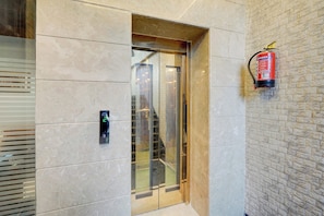Um elevador moderno com porta de vidro, extintor de incêndio fixado na parede e painel de exibição digital.