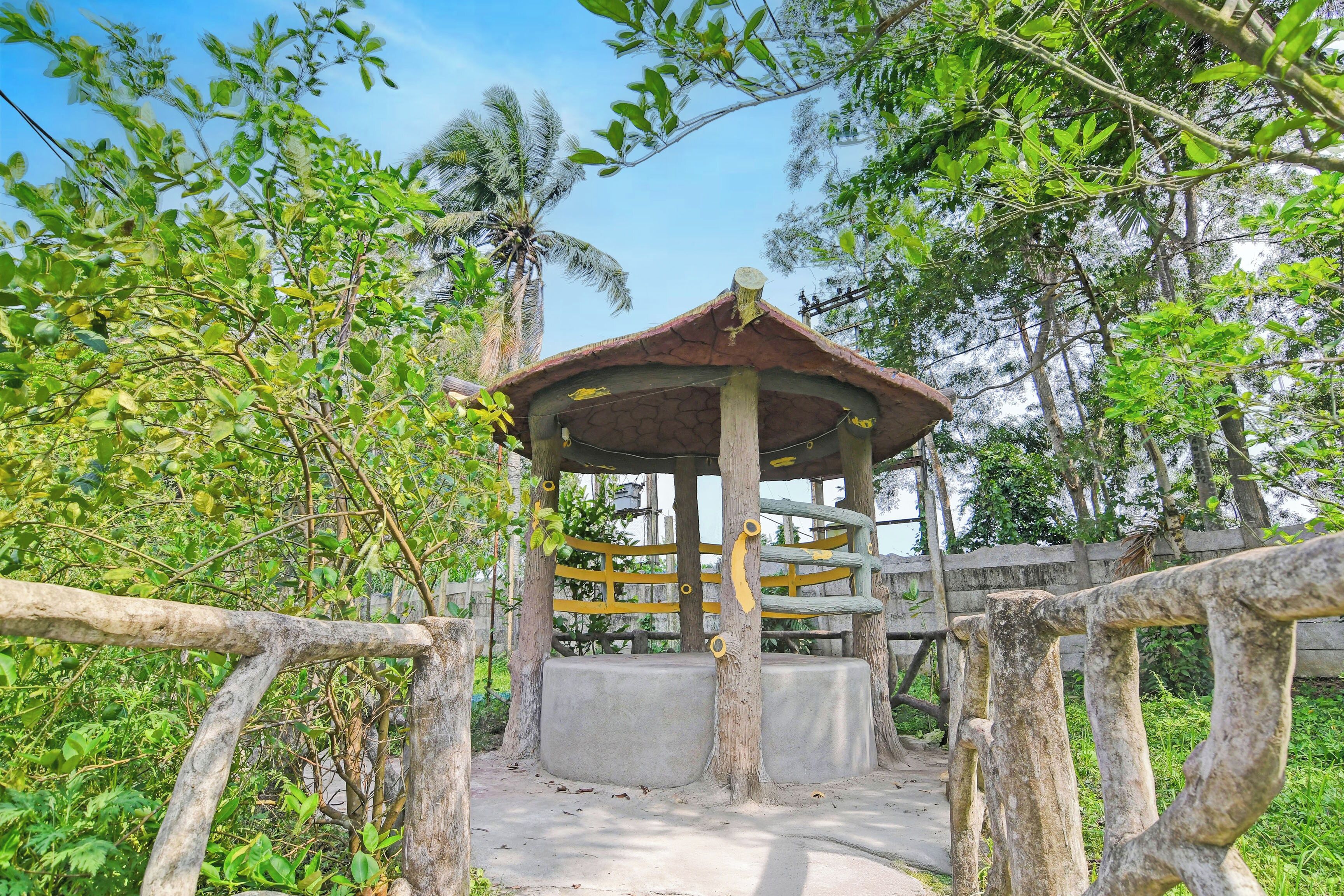 Um gazebo com telhado de madeira e base de concreto, cercado por uma cerca de madeira e vegetação exuberante.