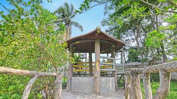 Um gazebo com telhado de madeira e base de concreto, cercado por uma cerca de madeira e vegetação exuberante.