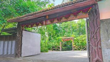 Uma entrada murada com um arco de madeira decorado com flores pendentes, que dá acesso a uma área pavimentada com uma parede de concreto e um portão de metal.