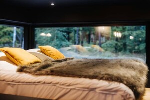 Uma cama com um cobertor fofinho e duas almofadas, uma janela com vista para árvores e uma luminária no teto.