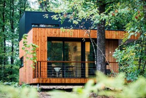 Uma cabana moderna de madeira com uma janela grande e varanda, rodeada por árvores.