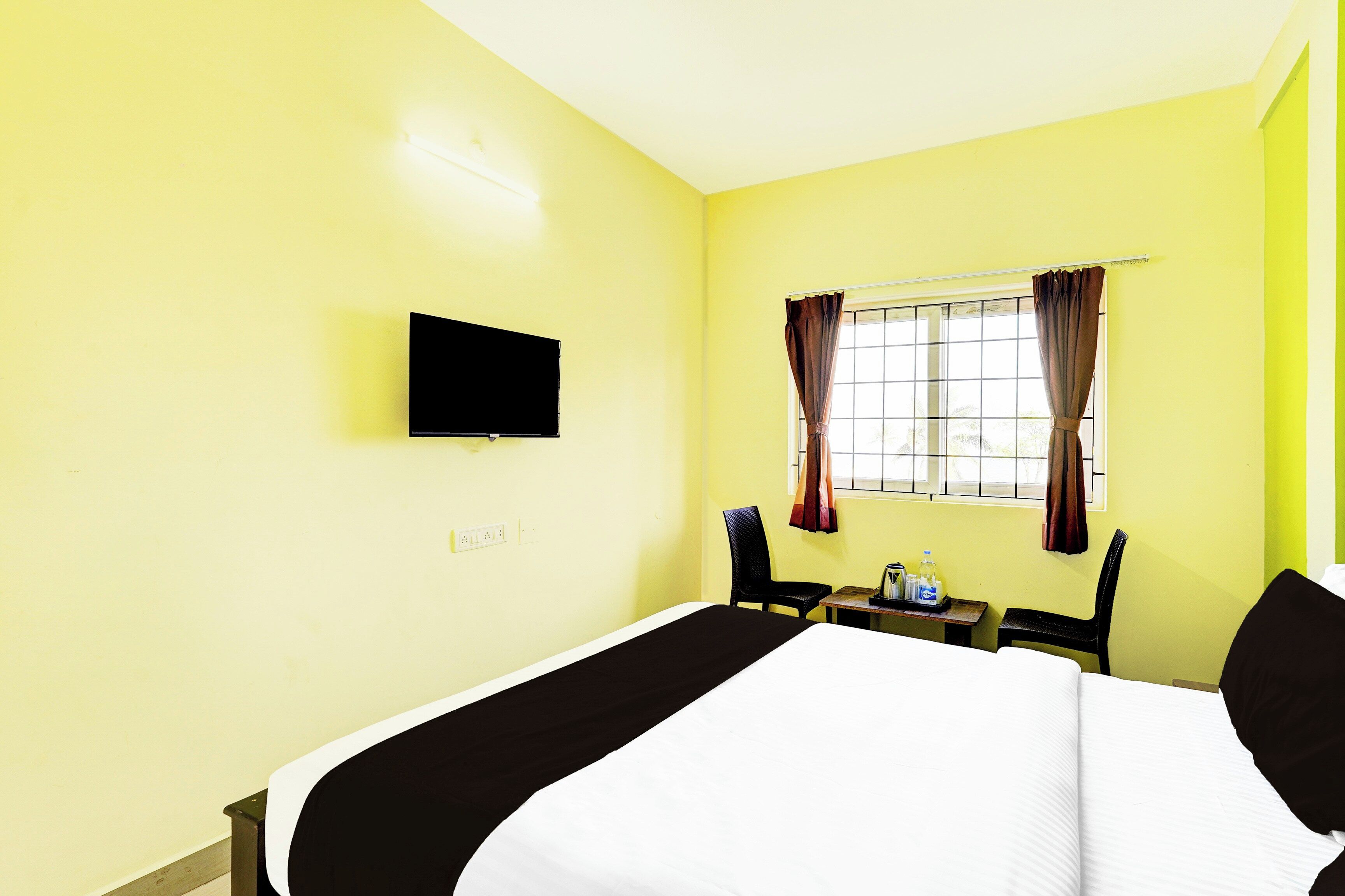 Quarto de hotel com cama, televisão na parede, janela com cortinas, mesa pequena com chaleira e garrafas, e uma cadeira.