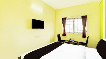 Quarto de hotel com cama, televisão na parede, janela com cortinas, mesa pequena com chaleira e garrafas, e uma cadeira.