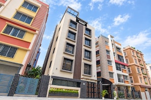 Um edifício residencial de vários andares com design moderno, destacando um elemento vertical marcante na fachada, entrada com portão e um céu claro ao fundo.