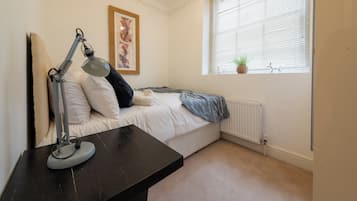 Quarto com cama, escrivaninha com luminária, radiador, janela com persianas e quadro na parede.