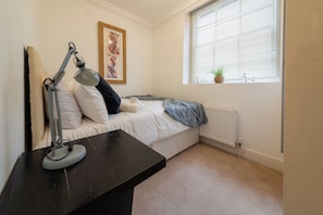 Quarto com cama, escrivaninha com luminária, radiador, janela com persianas e quadro na parede.