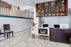 Uma área de recepção de hotel com piso de mármore, balcão de madeira, uma cadeira, um objeto decorativo na parede e uma varanda com corrimão.