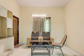 Sala de estar com sofá, mesa de centro e poltrona. Há uma janela com cortinas e uma estante com objetos decorativos.