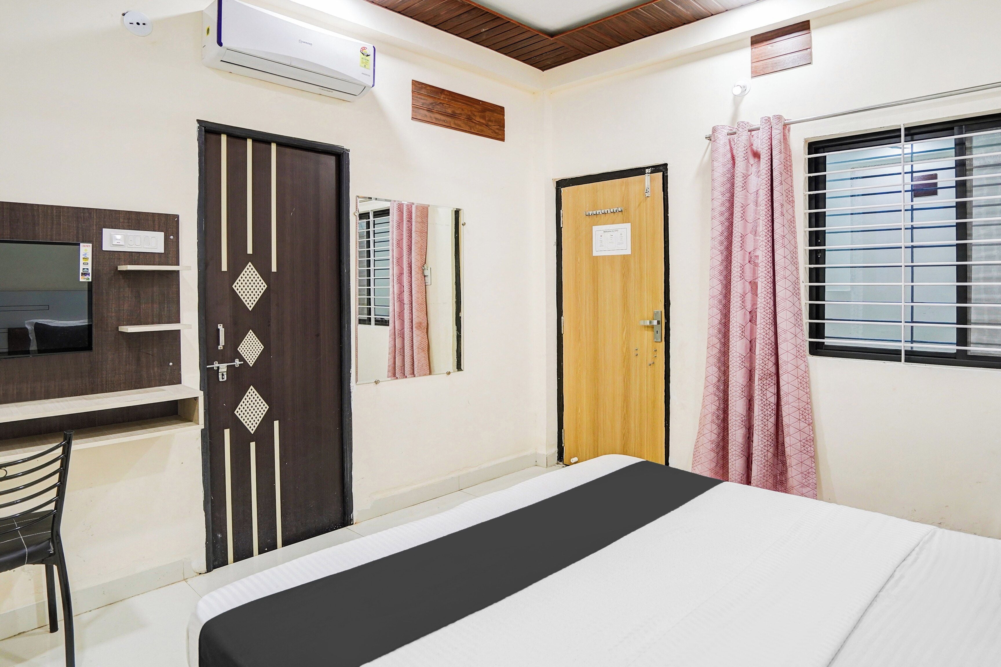 Quarto de hotel com cama, escrivaninha, cadeira, guarda-roupa, espelho, janela com cortinas e ar condicionado.