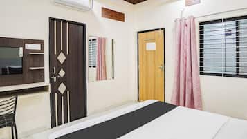 Quarto de hotel com cama, escrivaninha, cadeira, guarda-roupa, espelho, janela com cortinas e ar condicionado.