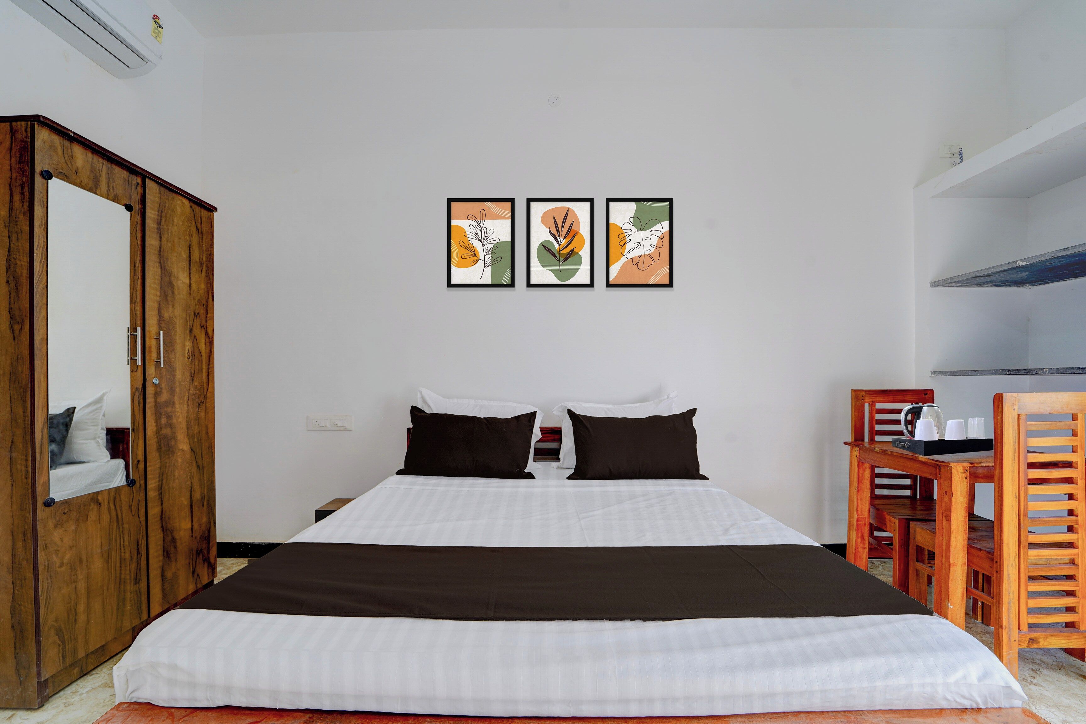 Quarto de hotel com uma cama, guarda-roupa de madeira, escrivaninha com cadeira e três obras de arte emolduradas na parede.