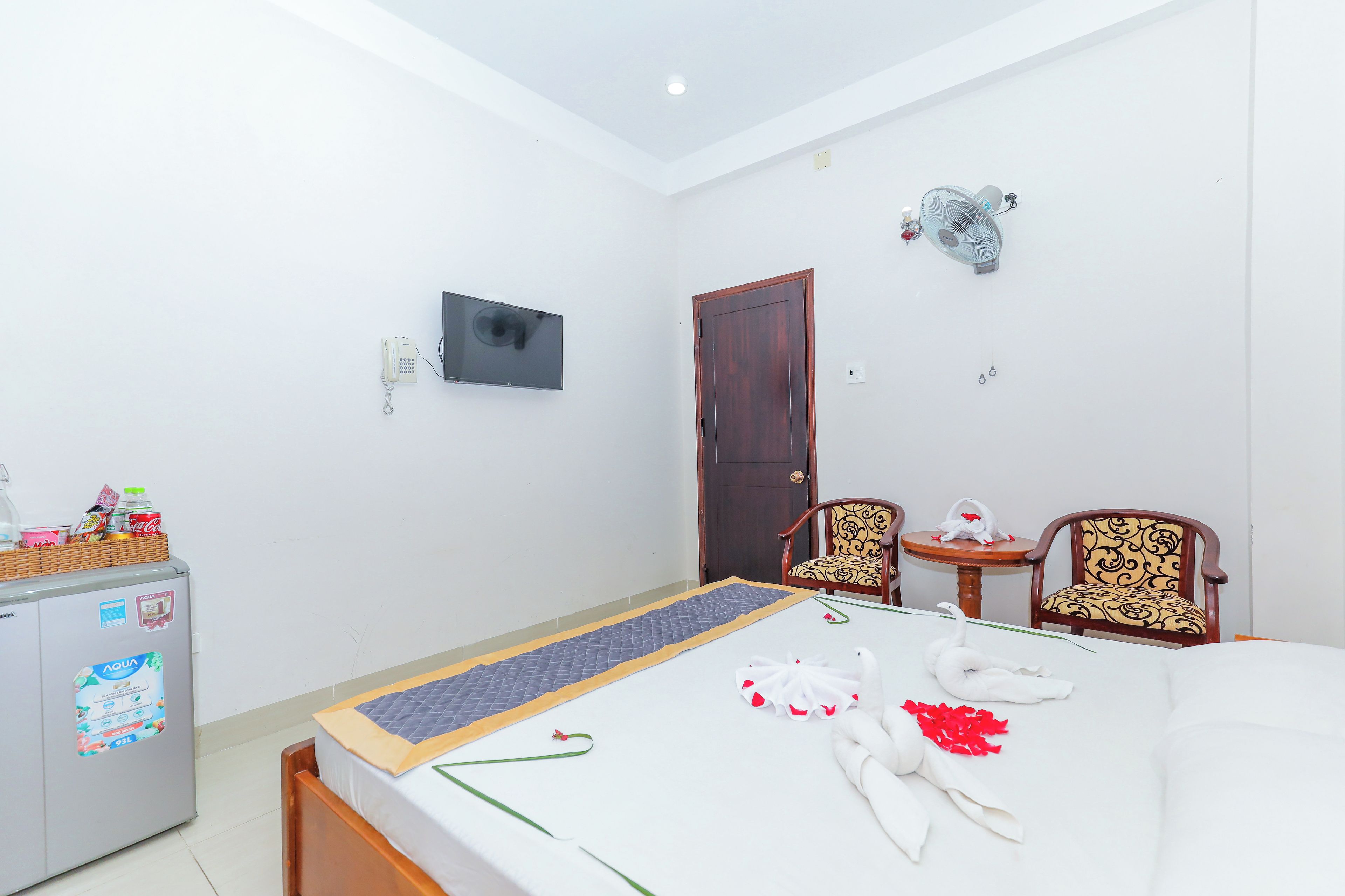 Quarto de hotel com cama, duas cadeiras, uma mesa pequena, televisão e frigorífico.
